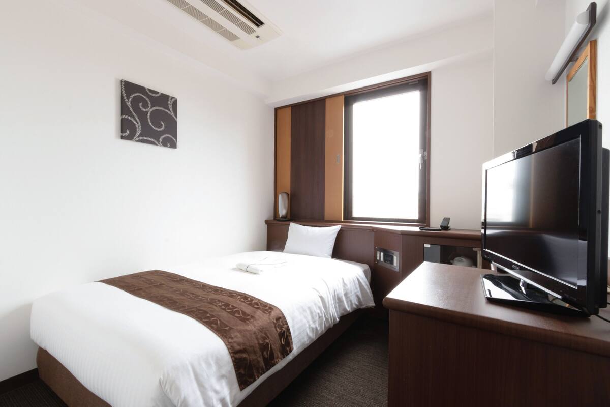Quarto de hotel com cama, escrivaninha com televisão de tela plana, janela e ar-condicionado fixado na parede.