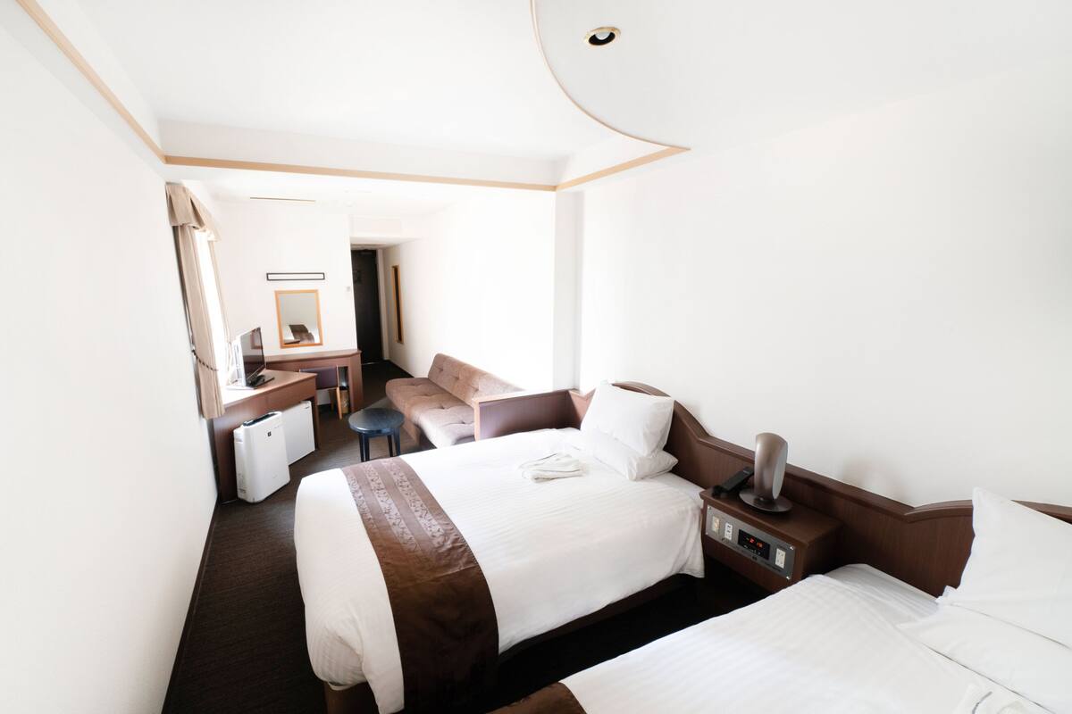 Quarto de hotel com duas camas, uma escrivaninha, uma cadeira, uma mesinha e um espelho.