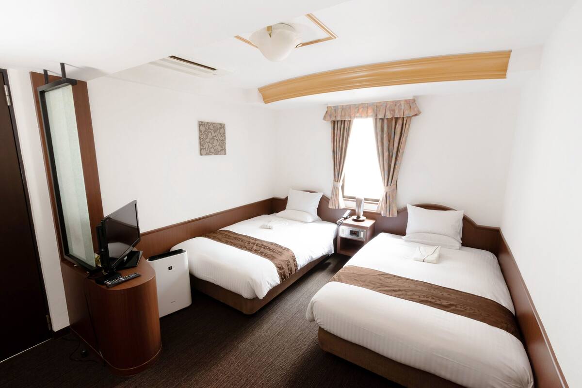 Quarto de hotel com duas camas, televisão, escrivaninha e janela com cortinas.