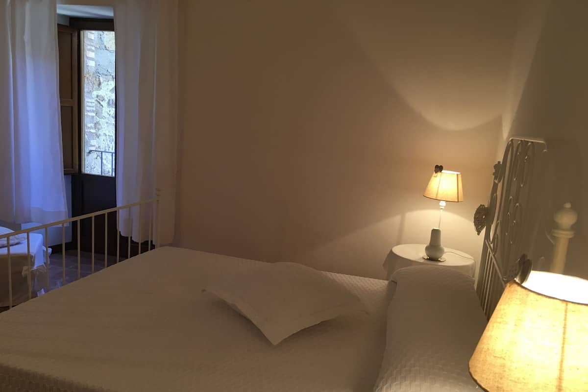Quarto com cama, mesa de cabeceira com abajur, janela com cortinas e vista para o exterior.