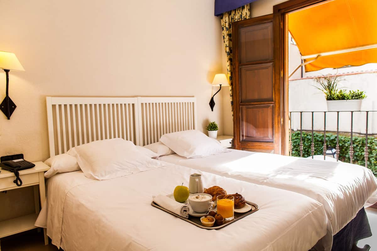 Cama bem arrumada com lençóis brancos, bandeja com itens de café da manhã incluindo um croissant, muffin e suco de laranja, e mesa de cabeceira com abajur e telefone.