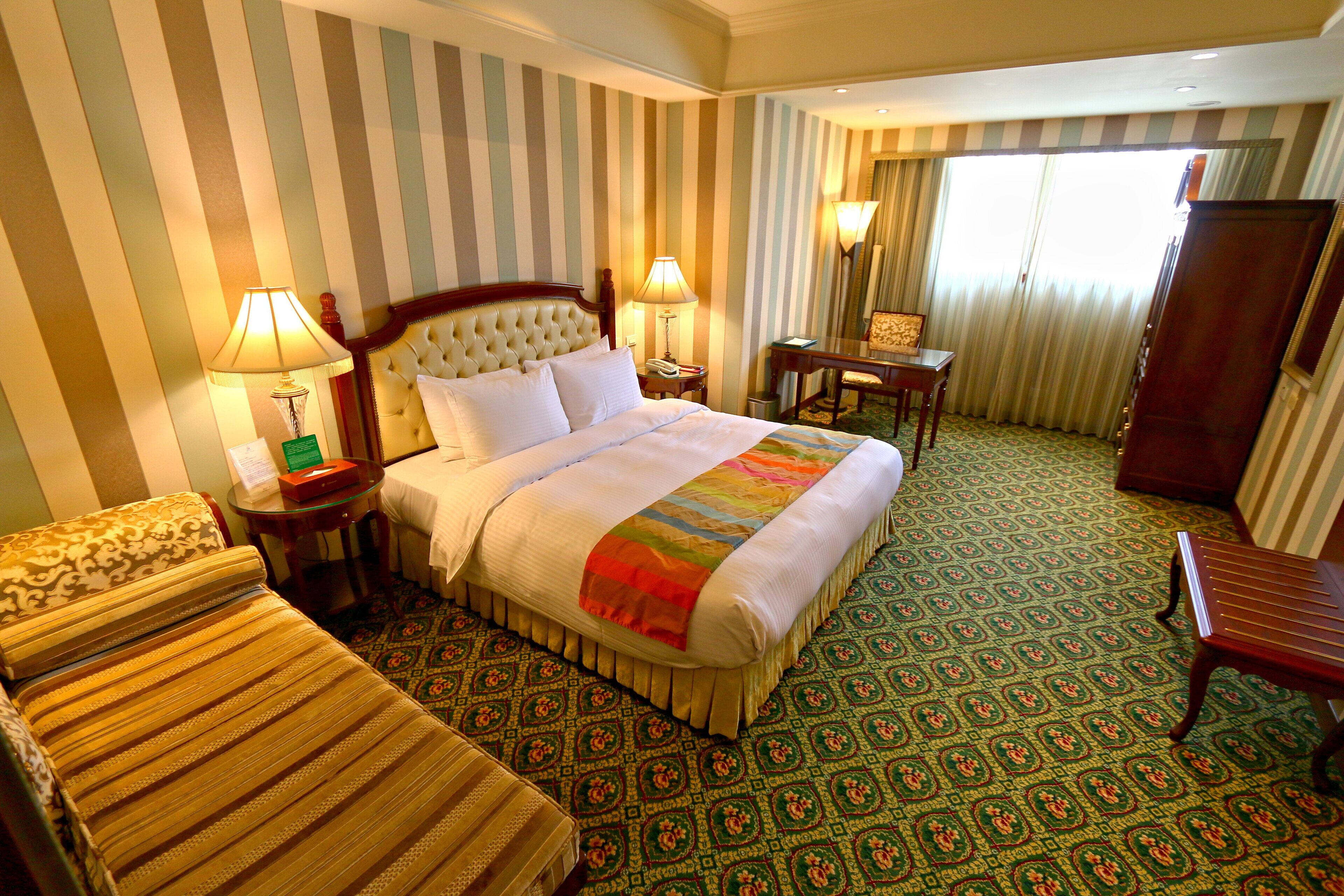 Quarto de hotel com uma cama grande, duas mesas de cabeceira com luminárias, uma cadeira, uma escrivaninha e um guarda-roupa.