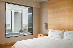 Quarto de hotel moderno com uma grande janela, cama com roupa de cama branca, mesinha de cabeceira e criado-mudo.