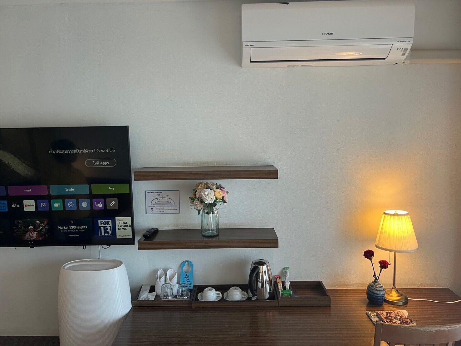 Quarto de hotel com televisão de tela plana exibindo interface webOS, ar-condicionado fixado na parede, prateleira de madeira com um vaso de flores, bandeja com produtos de higiene pessoal e luminária de mesa.