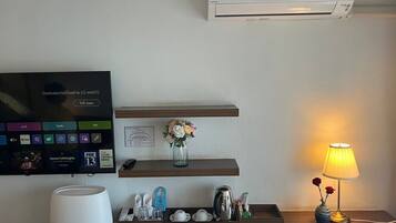 Quarto de hotel com televisão de tela plana exibindo interface webOS, ar-condicionado fixado na parede, prateleira de madeira com um vaso de flores, bandeja com produtos de higiene pessoal e luminária de mesa.