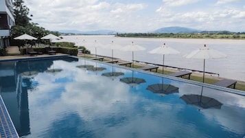 Piscina com espreguiçadeiras e guarda-sóis, com vista para um rio e montanhas ao longe.