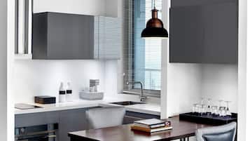 Uma cozinha moderna com armários cinza, uma coifa de aço inoxidável, uma janela com persianas, uma área de jantar com mesa e cadeiras, e uma luminária pendente.