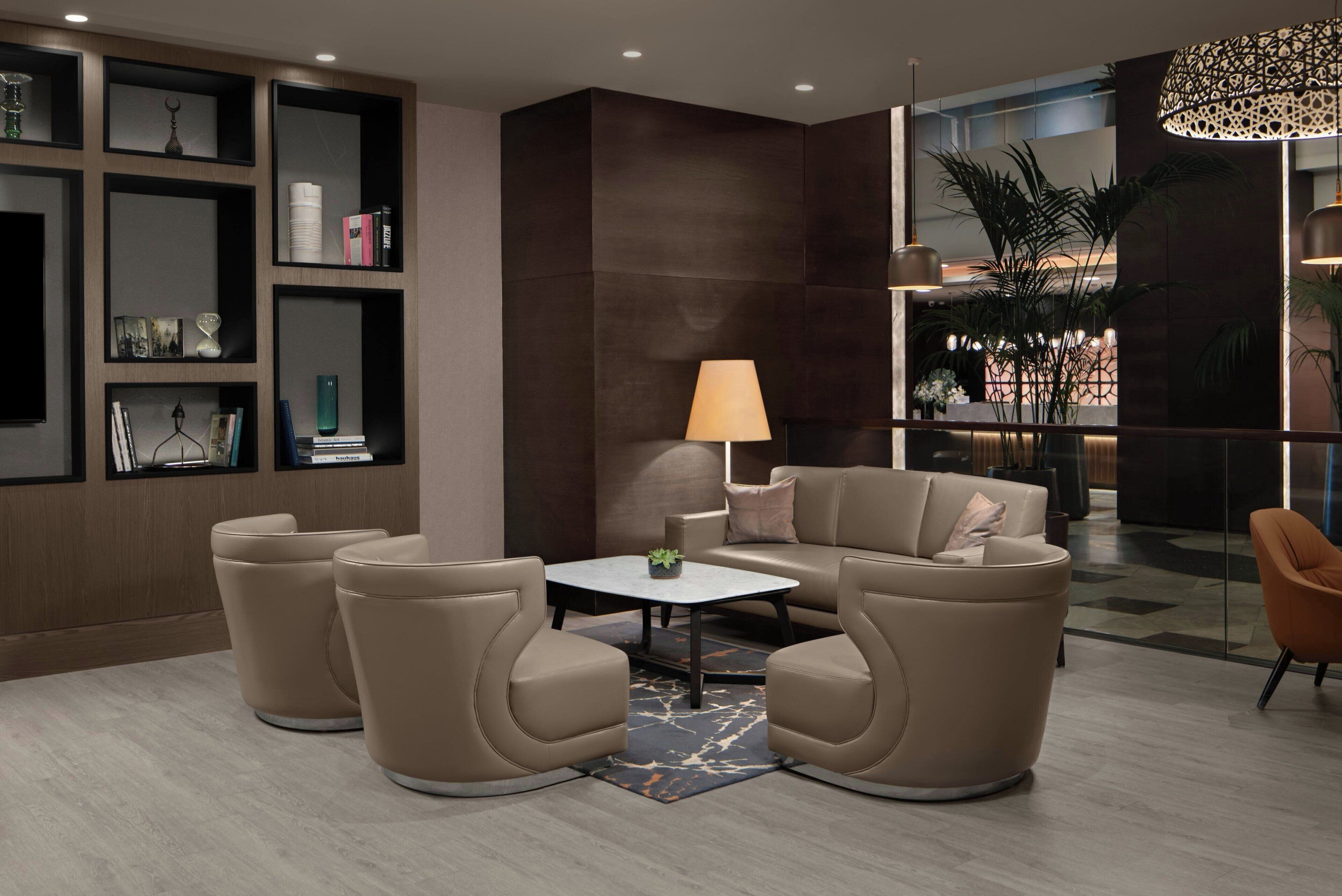 Uma área de estar moderna com um sofá, duas poltronas, uma mesa de centro e uma luminária. Há prateleiras com diversos objetos e uma janela grande com plantas.