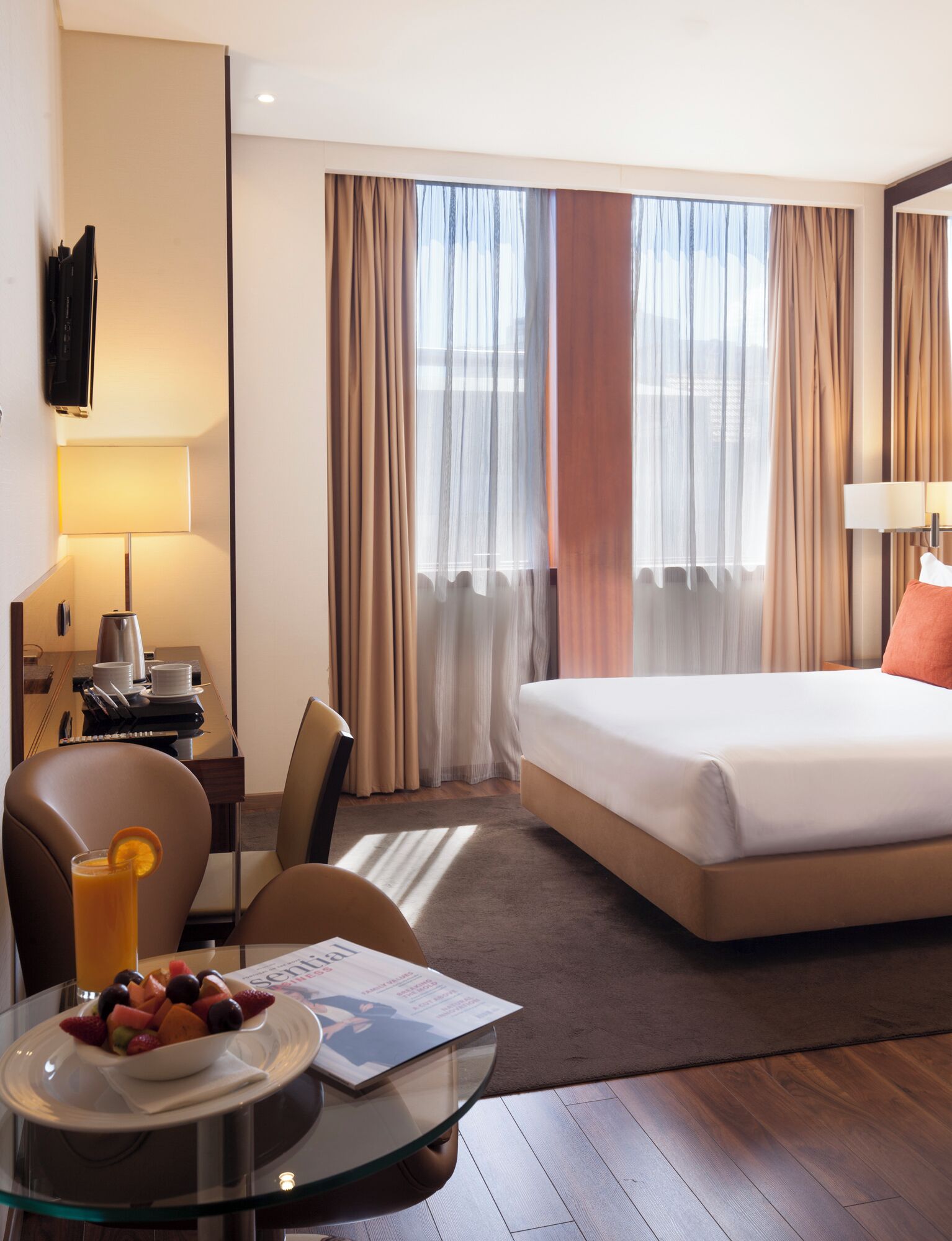 Quarto de hotel com cama, escrivaninha com luminária, cadeira, mesa de vidro com frutas e um menu, e janelas amplas com cortinas.