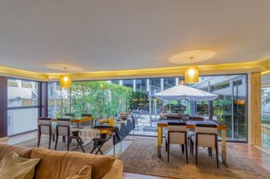 Uma área de refeições interna com mesas, cadeiras, um sofá e vista através de grandes janelas.