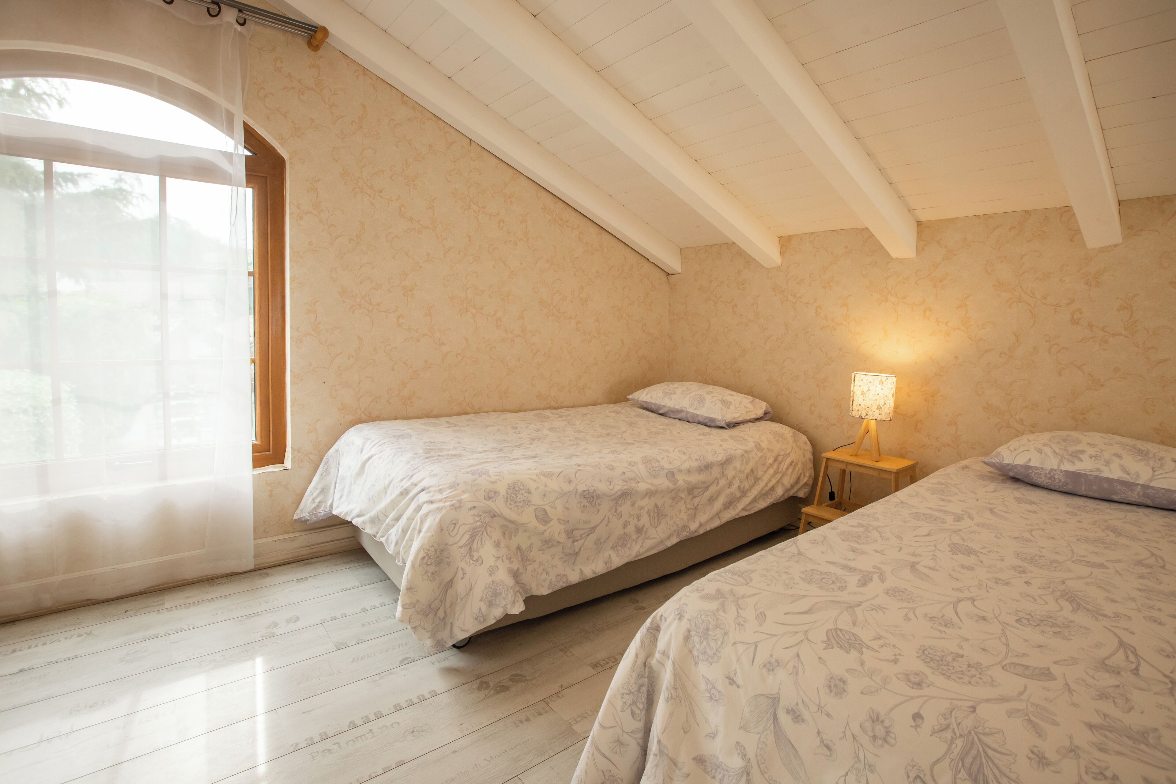 Quarto com duas camas, janela com cortina translúcida, luminária de cabeceira e teto de madeira.