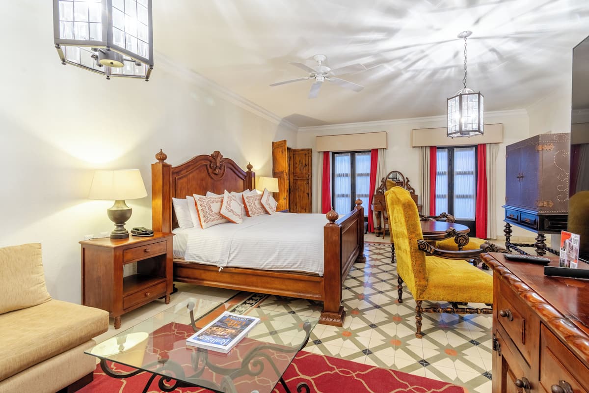 Quarto de hotel com cama de madeira, mesa de centro de vidro, poltrona amarela e cômoda de madeira.