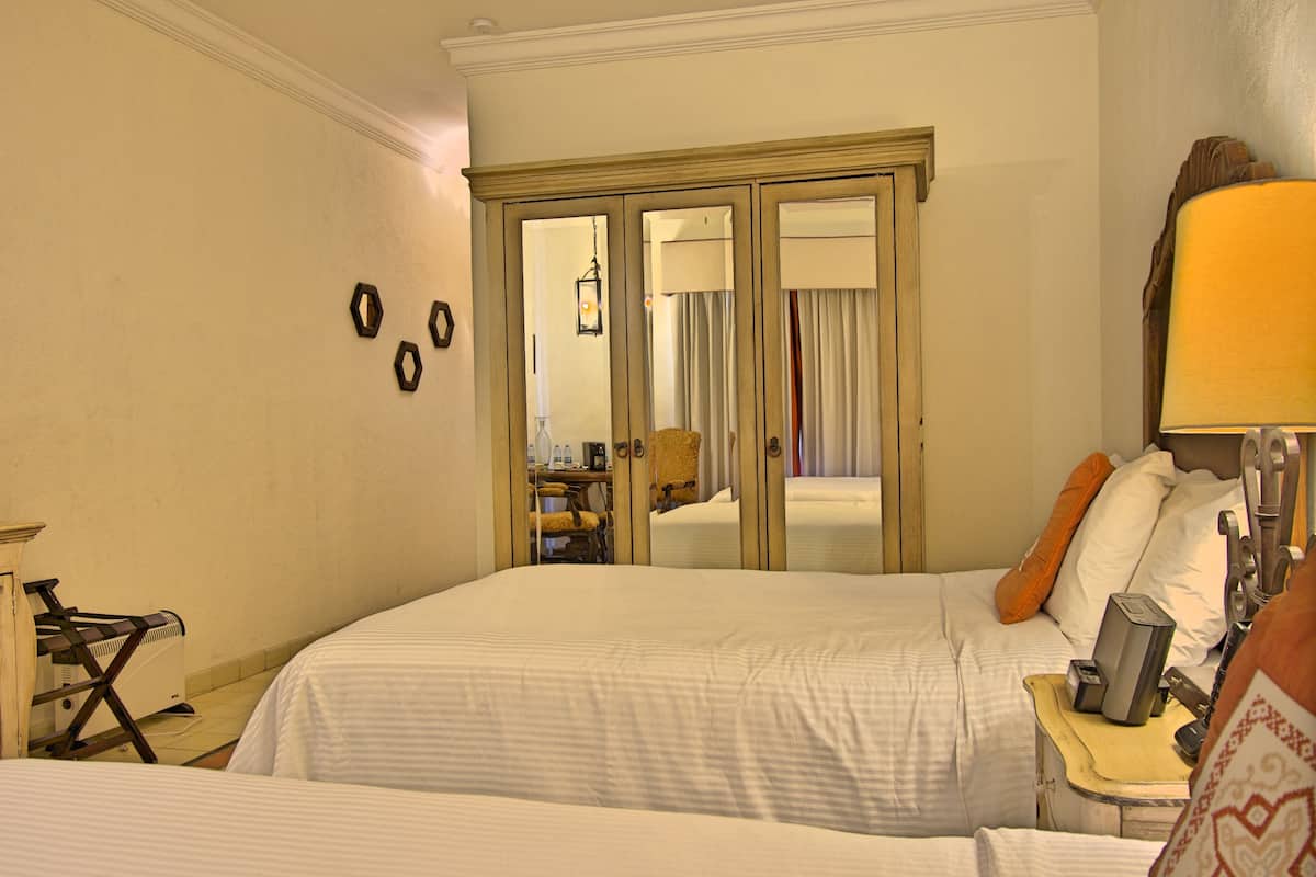 Quarto de hotel com duas camas, um guarda-roupa, uma escrivaninha com candeeiro e um espelho.