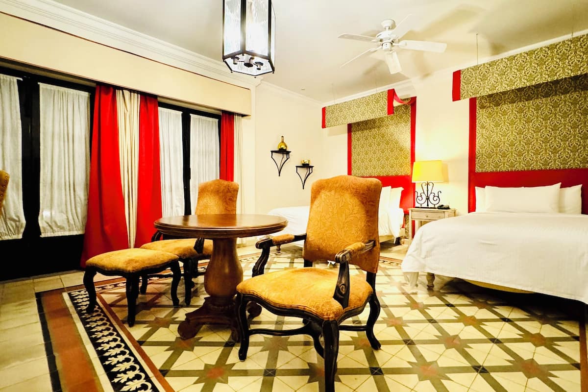 Quarto de hotel com cama, duas poltronas, uma mesa redonda, ventoinha no teto e parede estampada.