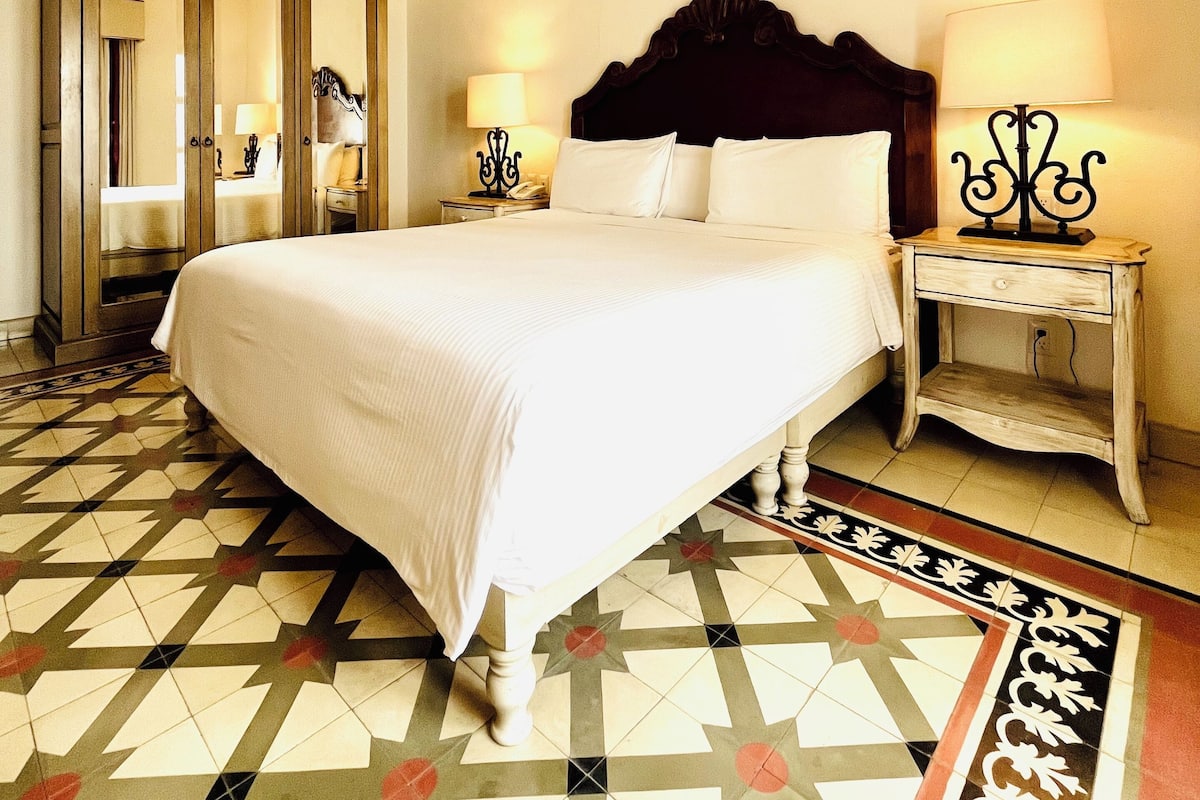 Quarto de hotel com uma cama grande, um ventilador de teto, uma mesinha de cabeceira com abajur, cômoda e espelho.
