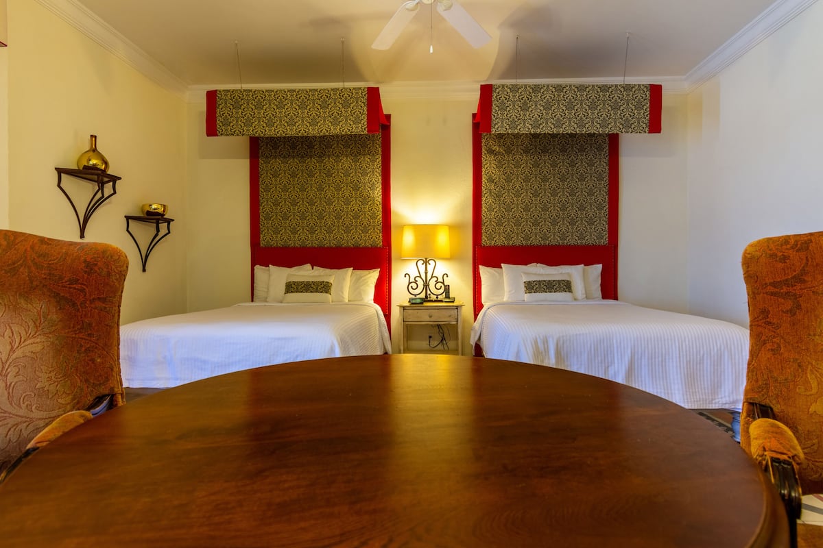 Quarto de hotel com duas camas de solteiro, uma mesa de madeira e duas poltronas.