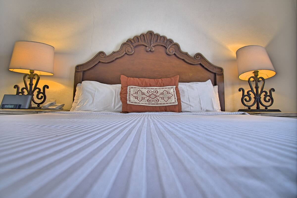 Cama bem arrumada com travesseiro estampado, duas luminárias de cabeceira e um telefone na mesinha de apoio.