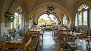 Uma ampla sala de jantar com altos tetos abobadados e grandes janelas, com mesas e cadeiras de madeira e um lustre central.