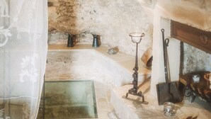 Uma casa de banho rústica de pedra com banheira, uma pequena janela e uma prateleira de madeira com um jarro e uma vela.