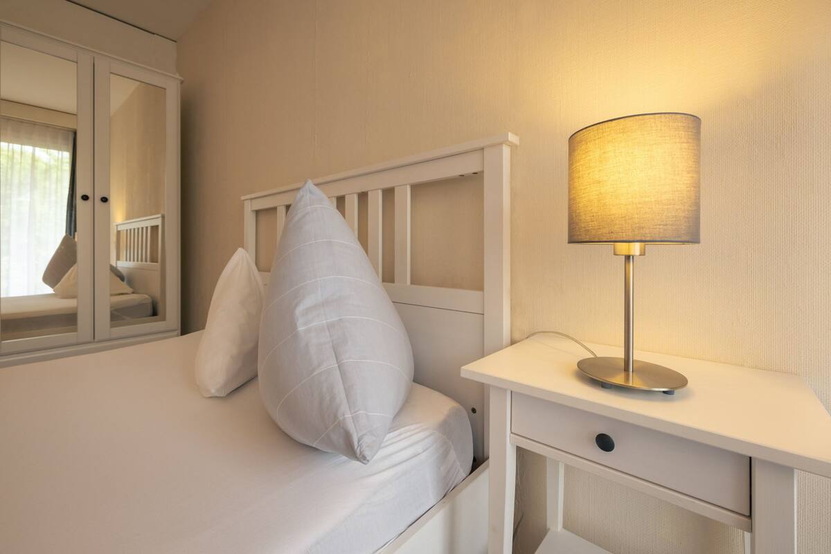 Uma cama bem arrumada com roupa de cama e almofadas brancas, uma mesa de cabeceira com abajur e um espelho na parede.