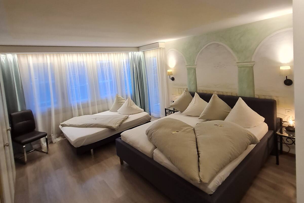 Quarto de hotel com uma cama grande, duas mesinhas de cabeceira, uma cadeira e vista da cidade através de cortinas translúcidas.