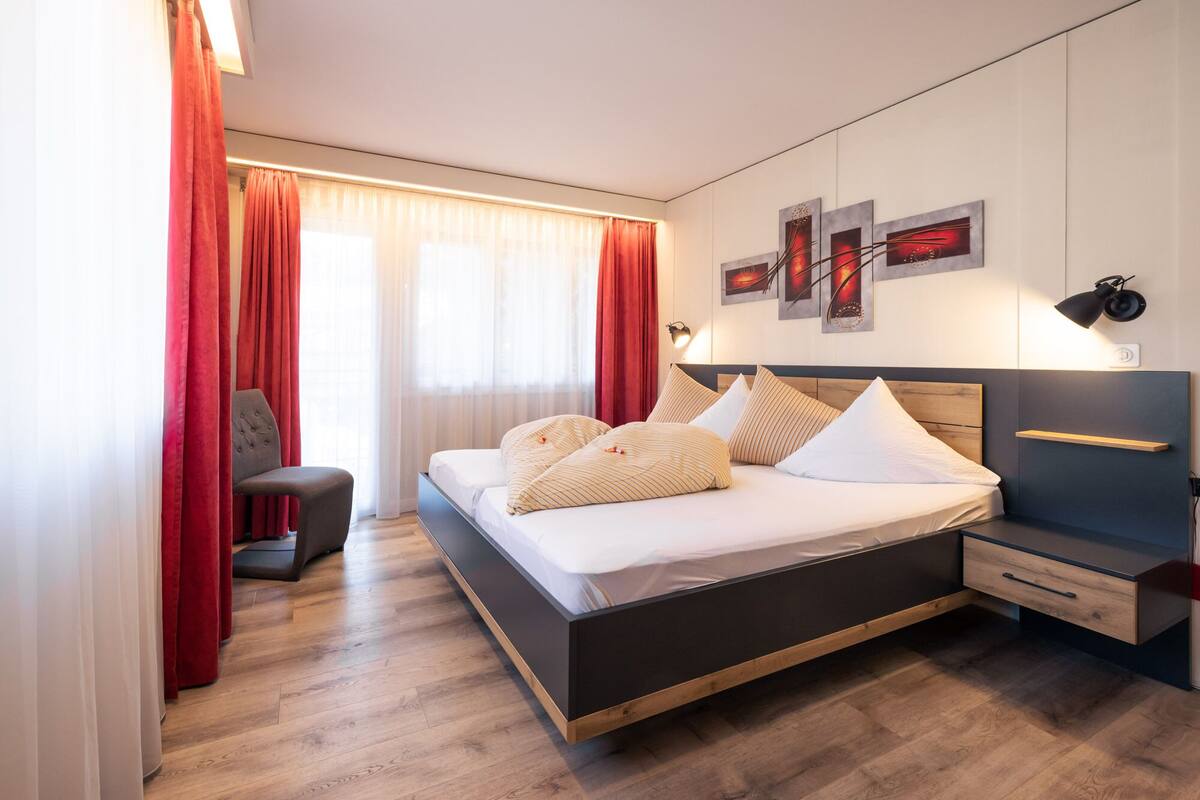 Quarto de hotel moderno com uma cama grande, uma mesinha de cabeceira, uma cadeira e uma janela com cortinas.