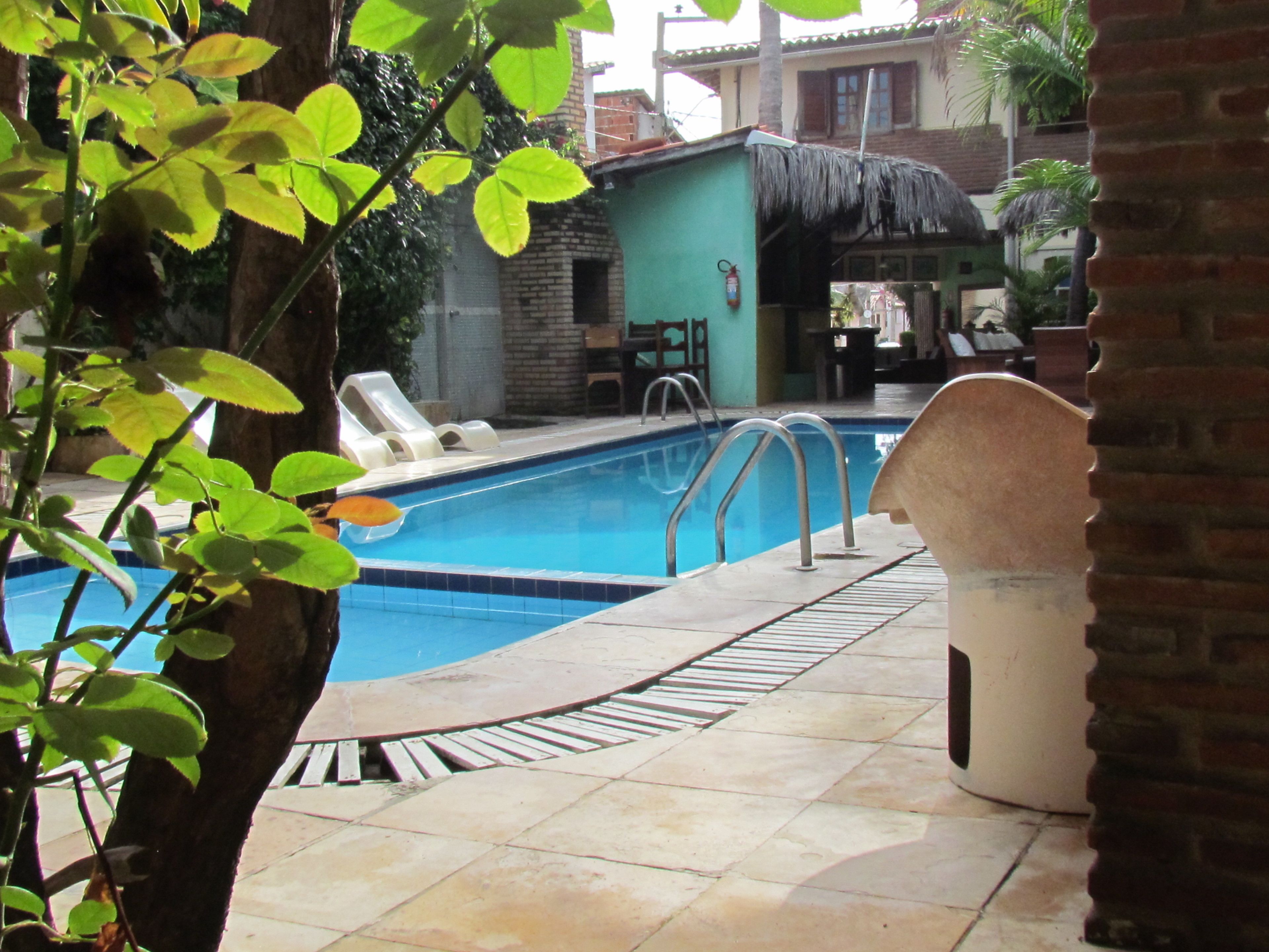 Uma área de piscina com um deck revestido de azulejos, cadeiras de descanso e um edifício de tijolos ao fundo.