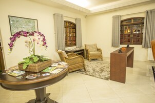 Quarto com uma mesa redonda exibindo revistas, uma cadeira de vime, uma pequena mesa de madeira com um objeto decorativo e uma janela com cortinas.