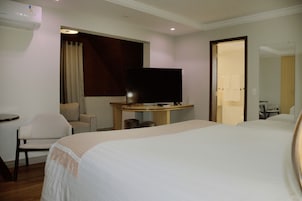 Quarto de hotel com uma cama grande, uma televisão em um suporte de madeira, uma escrivaninha com uma cadeira e um banheiro visível por uma porta aberta.