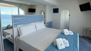 Quarto de hotel moderno com uma cama grande, vista para o mar, televisão de tela plana e uma mesa pequena.