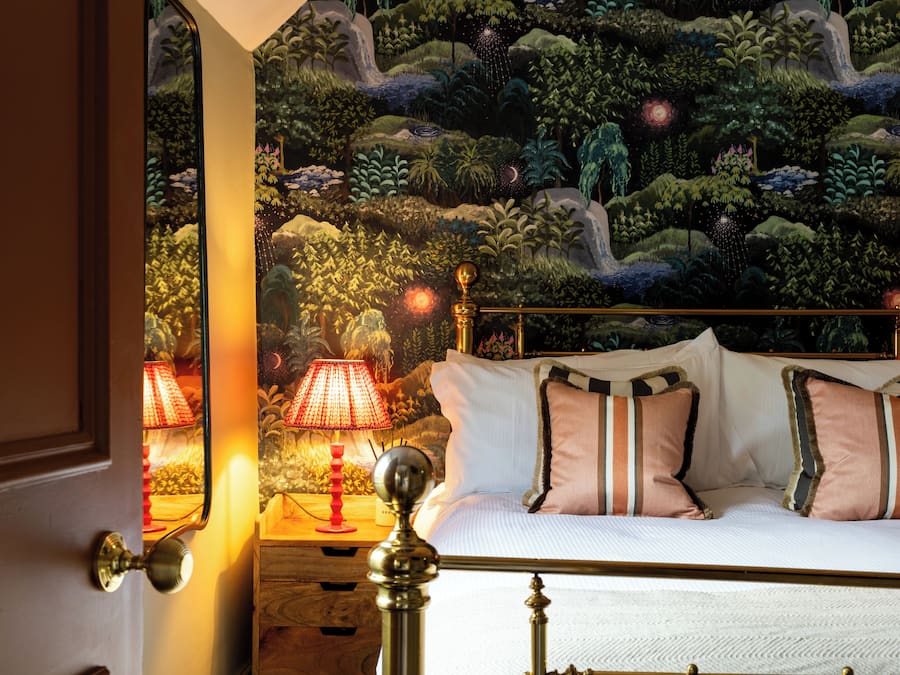 Quarto com cama, almofadas, mesa de cabeceira, abajur e um mural na parede.