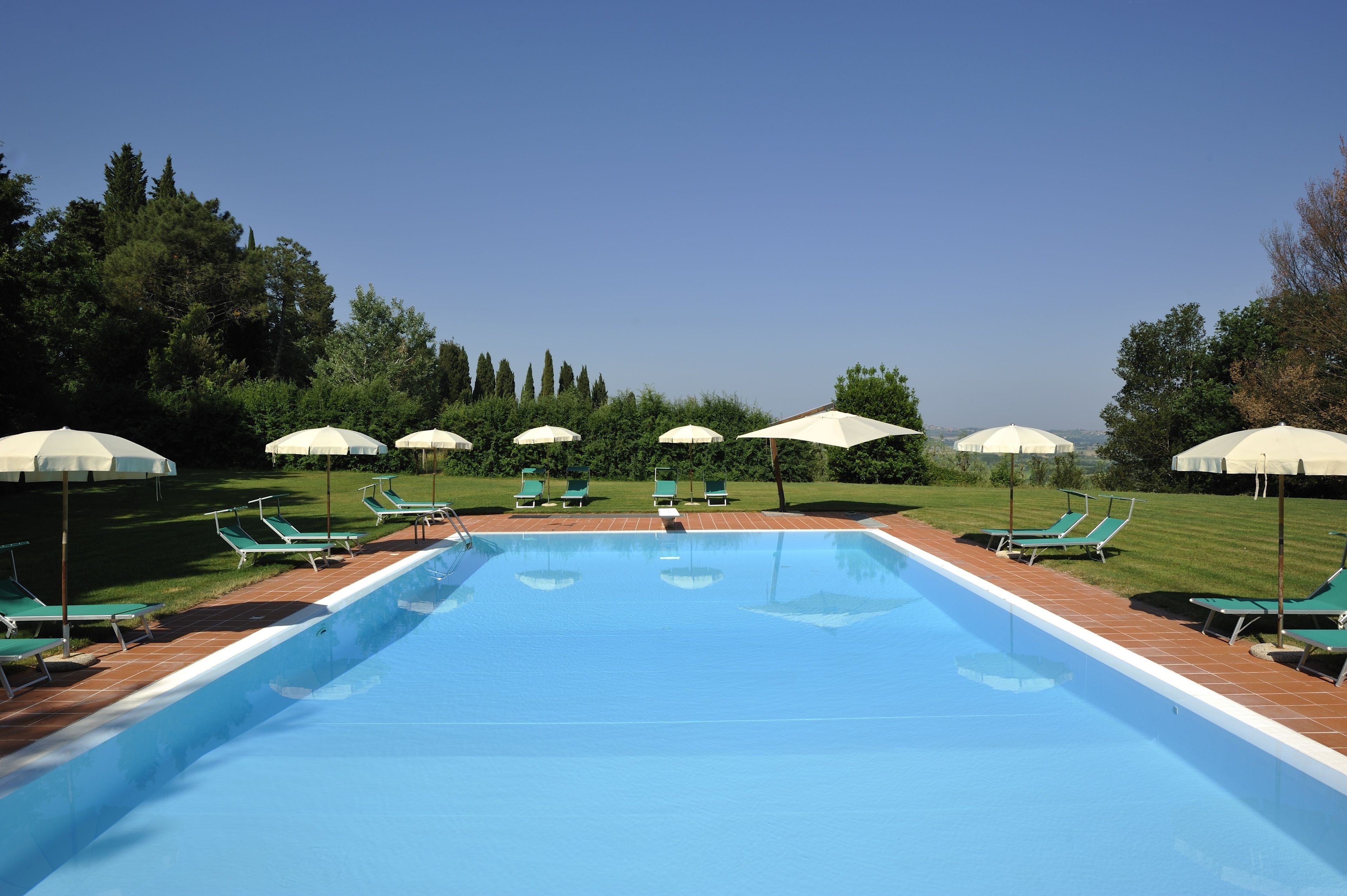 Piscina retangular com borda revestida, rodeada por cadeiras de grama verde e guarda-sóis brancos.