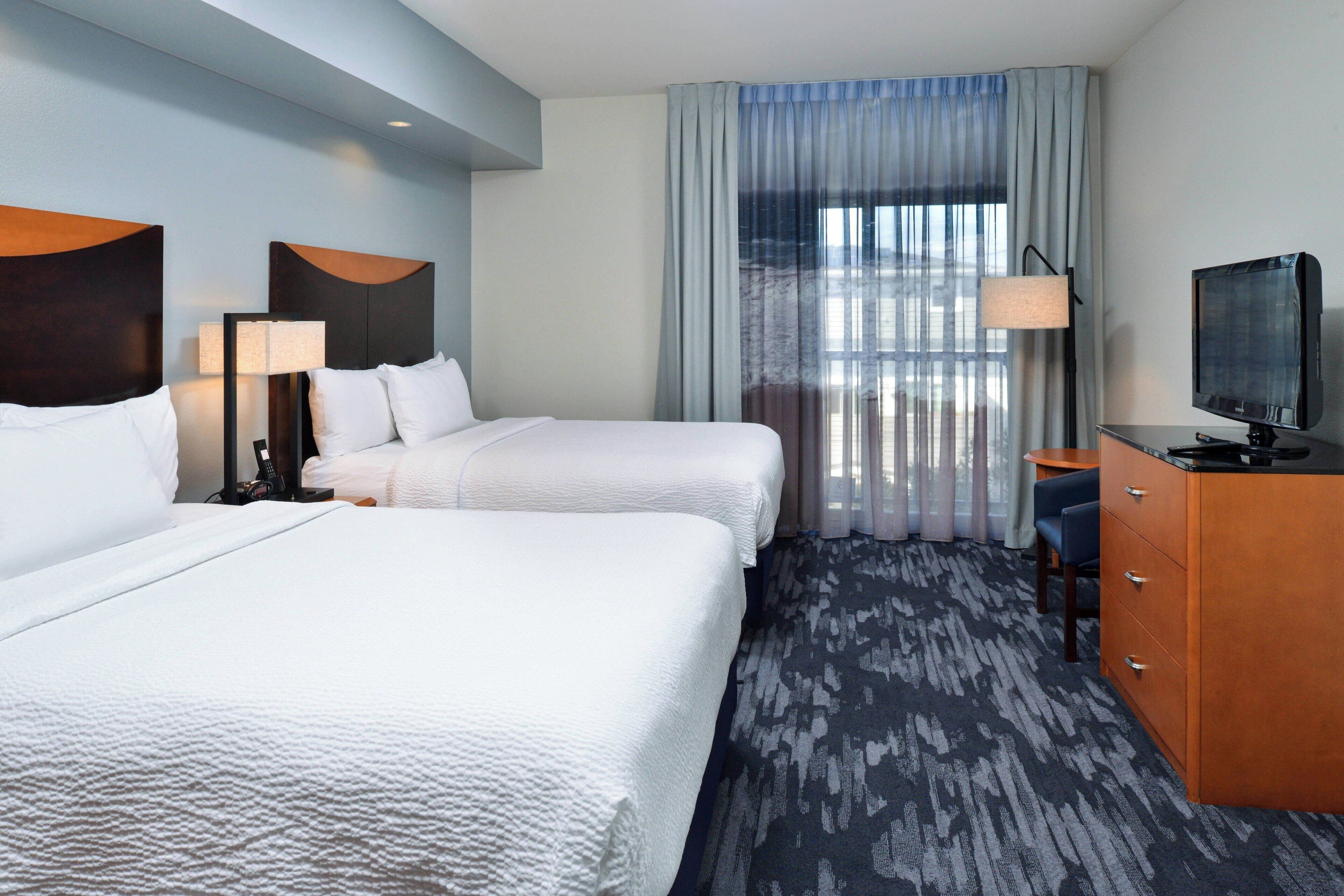 Quarto de hotel com duas camas, uma escrivaninha, televisão e uma janela grande com cortinas.