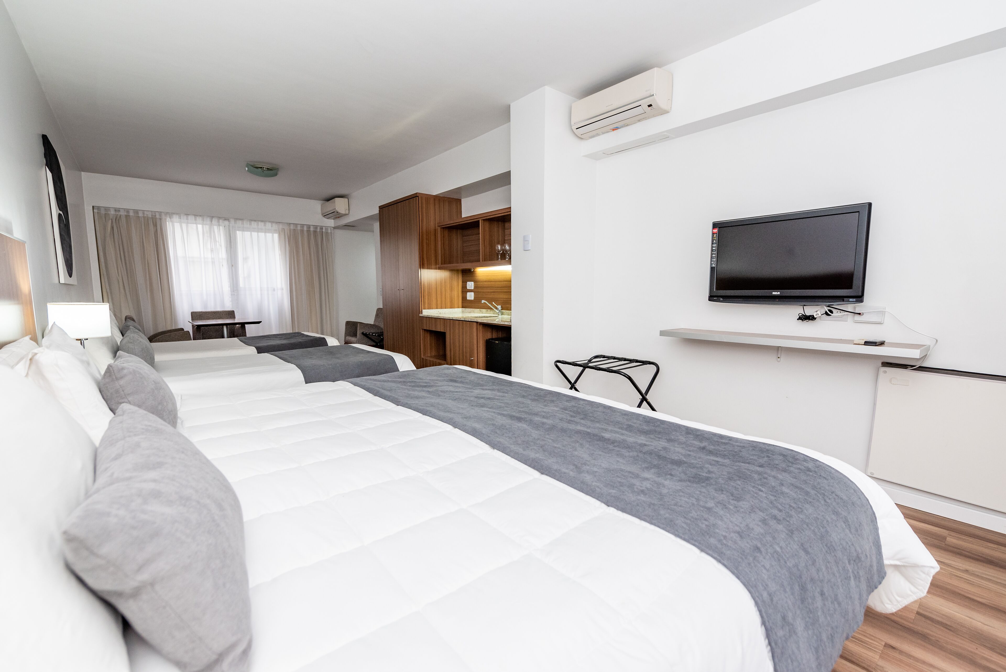 Quarto de hotel com uma cama grande, televisão montada na parede, pequena cozinha e janela com cortinas.