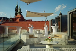 Terraço no telhado com área de estar externa, uma mesa com um vaso e vista para uma igreja com telhado vermelho e torres verdes.