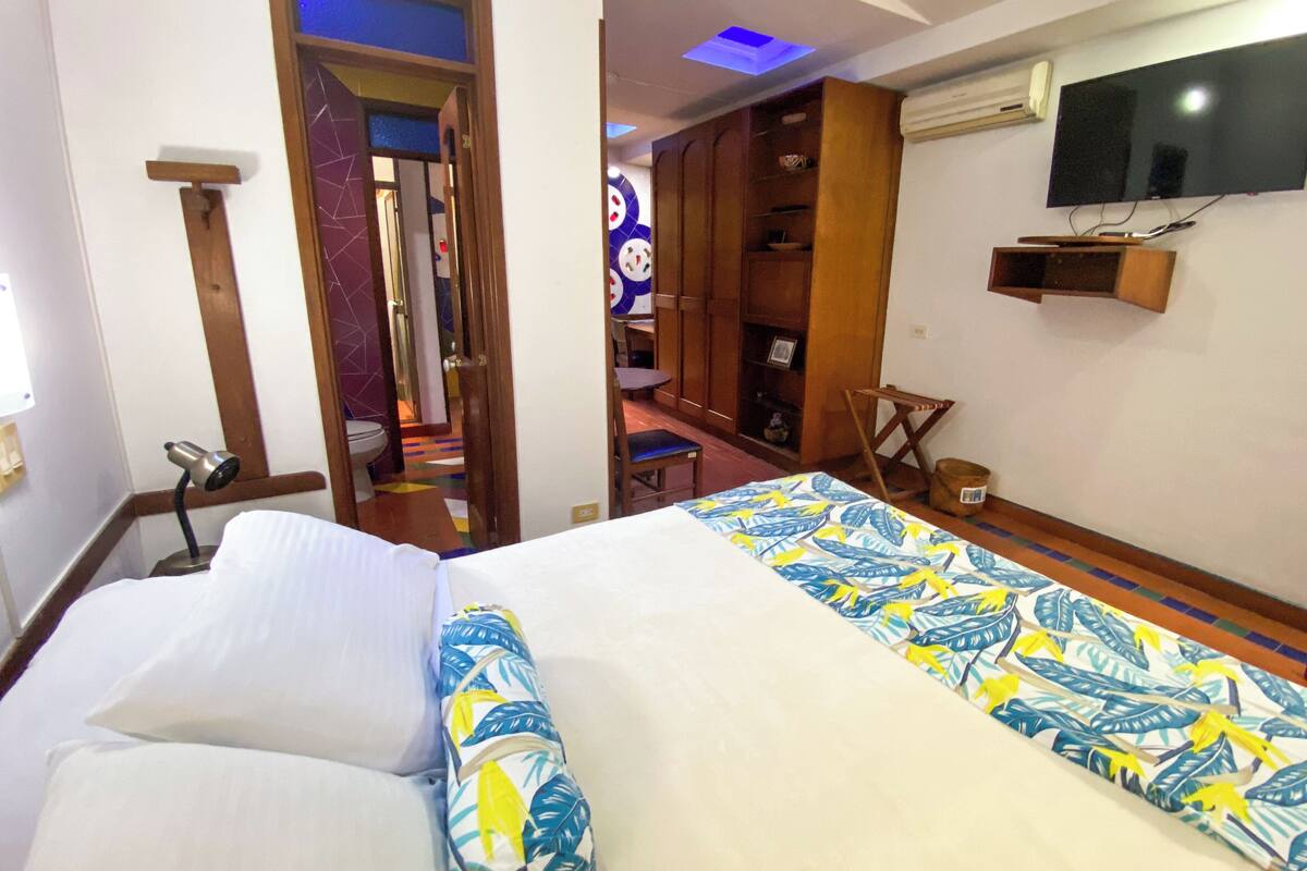 Quarto de hotel com cama, escrivaninha, cadeira, TV fixada na parede e banheiro visível por uma porta aberta.