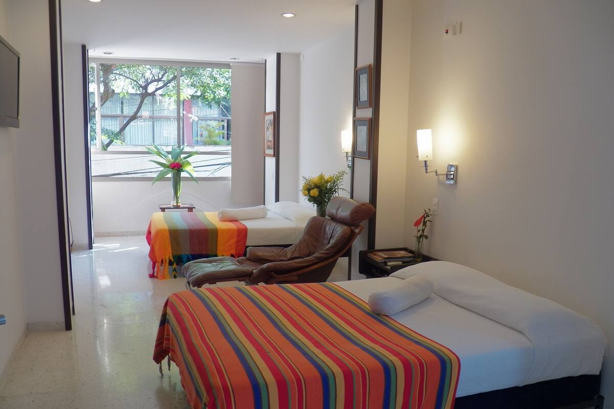 Quarto de hotel com cama, cadeira, mesinha e janela com vista para o exterior.