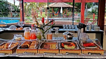 Um buffet de café da manhã com diversos alimentos e bebidas, piscina ao fundo, e vista para um jardim e área de estar.