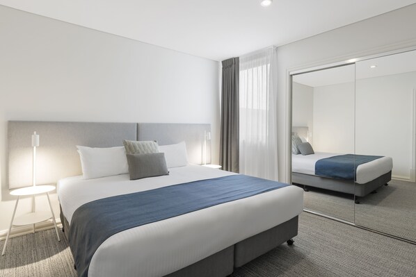 Quarto de hotel moderno com uma cama grande, uma mesa de cabeceira, um espelho e vista para o quarto vizinho.