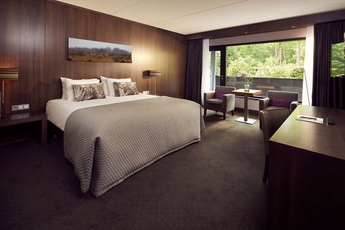 Quarto de hotel com uma cama grande, uma escrivaninha, uma cadeira, uma mesinha de centro e vista para o verde pela janela.