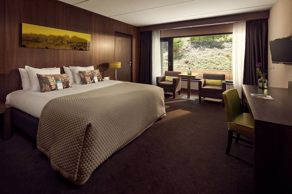 Quarto de hotel com uma cama grande, uma escrivaninha, duas cadeiras, uma mesa pequena e uma janela ampla com vista para vegetação.