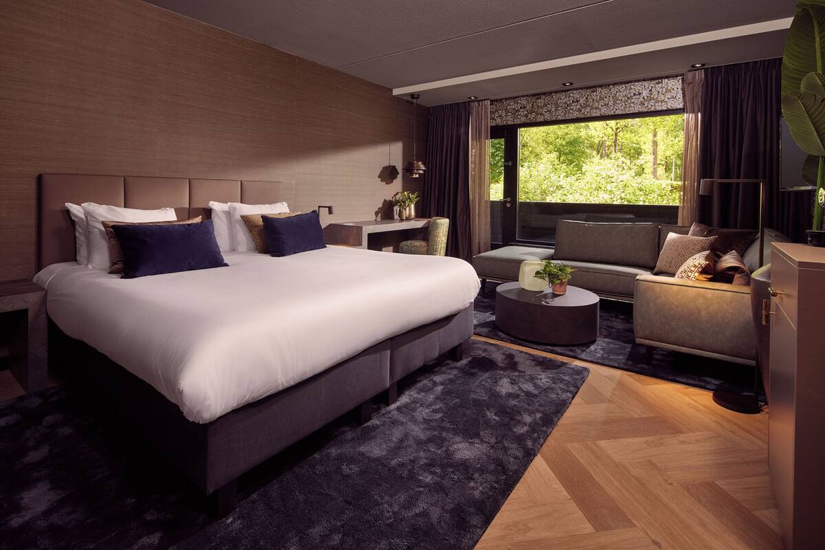 Quarto moderno com uma cama grande, um sofá, uma mesinha e vista para o verde através de uma janela.