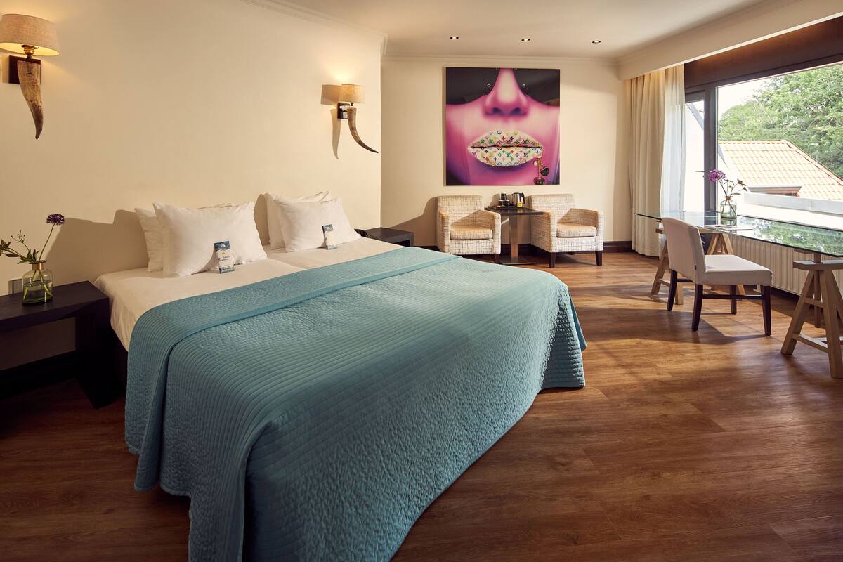 Quarto de hotel com uma cama grande, um espaço de estar com cadeiras, uma pequena mesa e um quadro na parede.