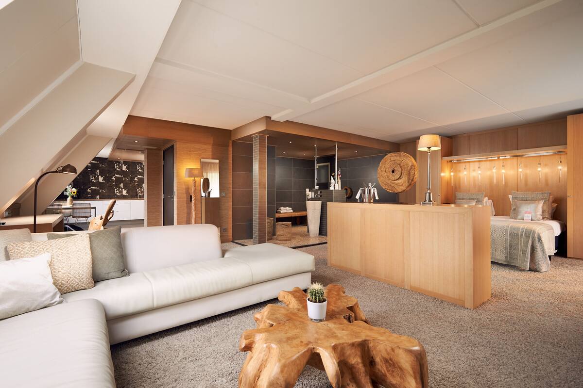 Quarto de hotel moderno com sofá, mesa de centro de madeira e cama com cabeceira.