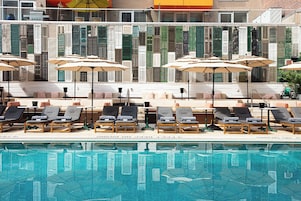 Uma área à beira da piscina com espreguiçadeiras e guarda-sóis, uma piscina em mosaico e um edifício com persianas ao fundo.