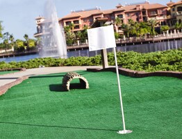 Um campo de mini golfe com uma fonte de água ao fundo e uma fileira de edifícios.