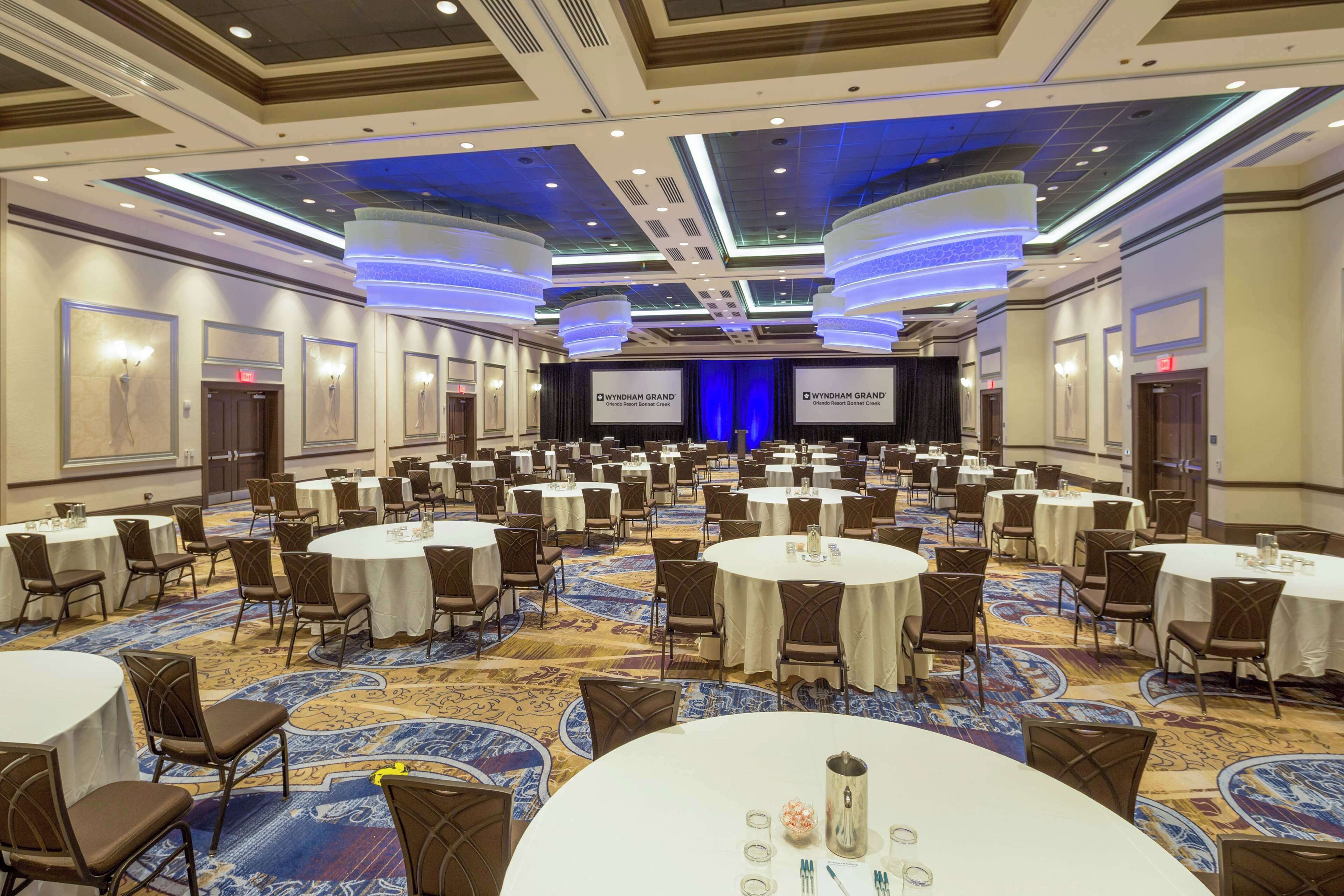 Uma grande sala de conferências com mesas e cadeiras redondas, um palco com fundo azul e piso coberto por carpete.
