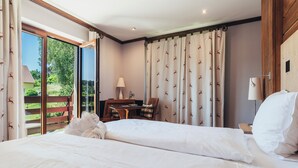 Quarto de hotel com uma cama grande, uma mesa com cadeira, uma luminária e vista para o verde lá fora.