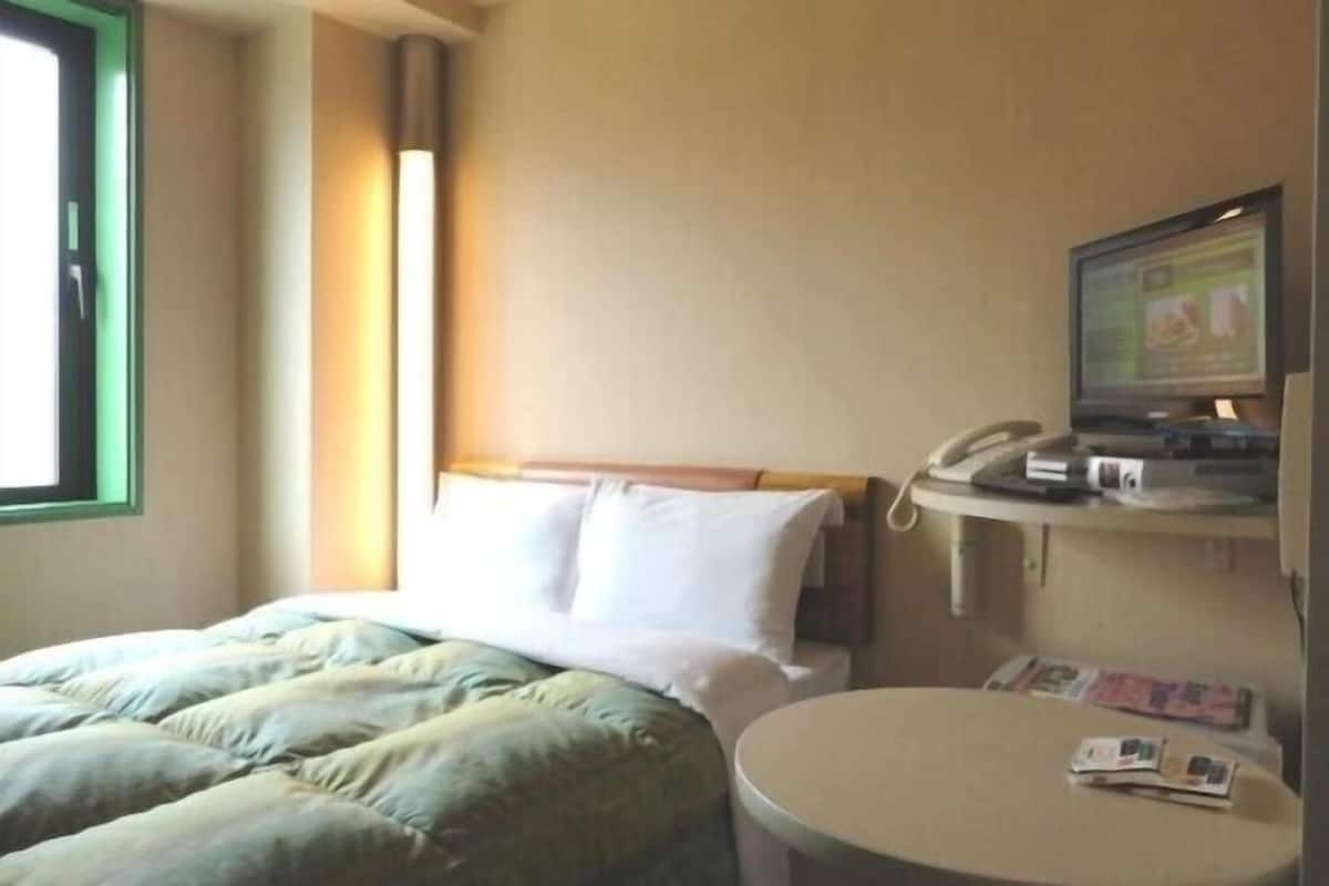 Quarto de hotel com uma cama, uma mesa redonda com uma revista, uma televisão e um telefone.