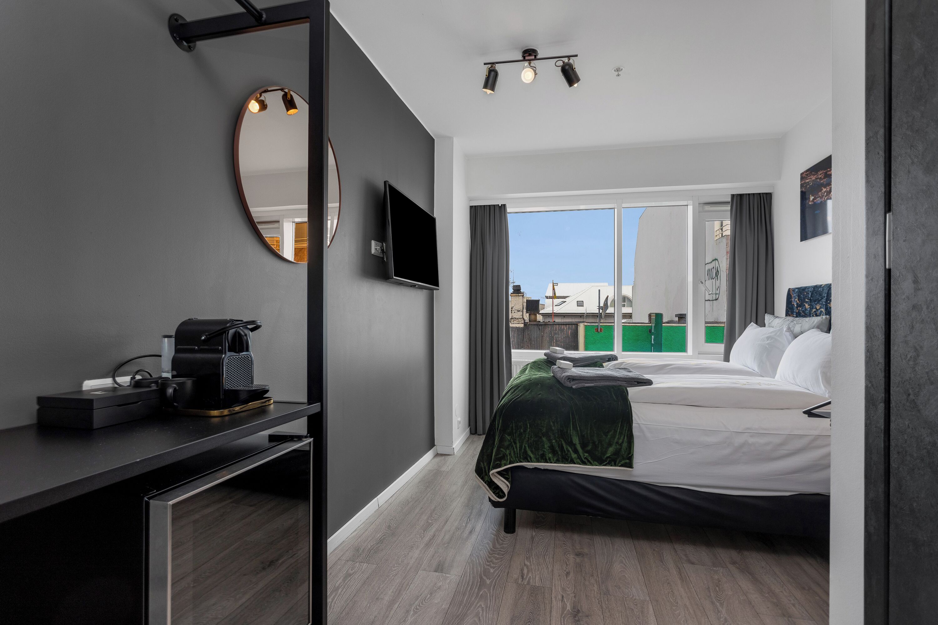 Quarto de hotel moderno com cama, escrivaninha com cafeteira, TV de tela plana e uma grande janela com vista para a cidade.